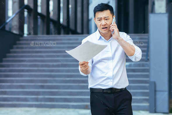 Frustrated and angry Asian businessman talking on the phone, holding a ...