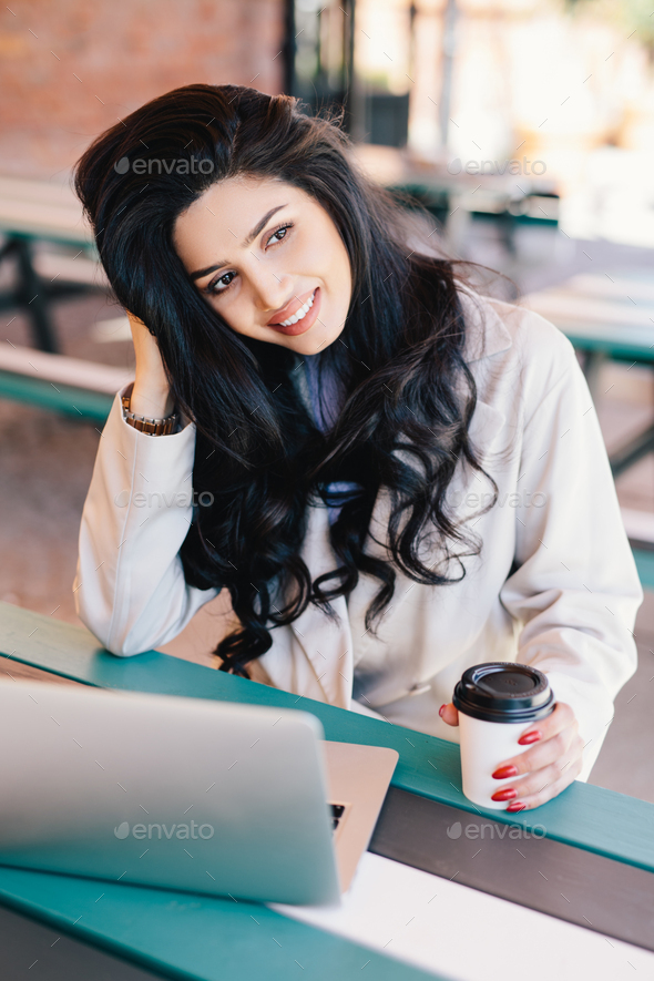 female with dark hair holding takeaway coffee sitting in front of ...
