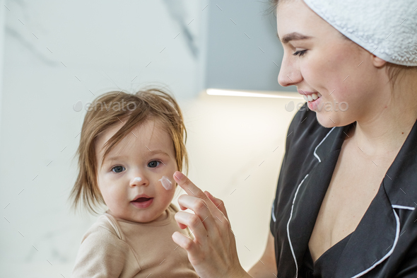 Mom puts baby cream on face of toddler daughter. Cozy house. Quality ...