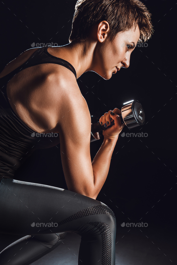 side view of female athlete exercising with dumbbell at gym, black ...