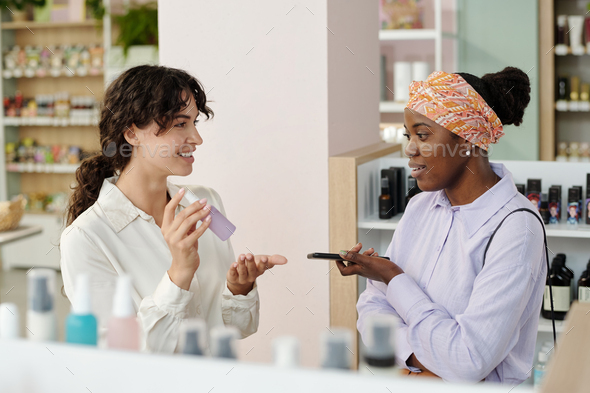 Young smiling shop assistant recommending black woman cosmetic product ...