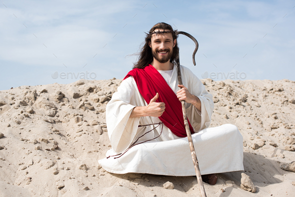smiling Jesus sitting in lotus position on sand, holding staff and ...