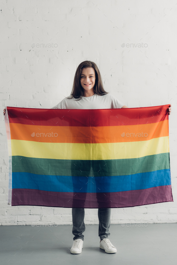 happy transgender woman holding pride flag in front of white brick wall ...