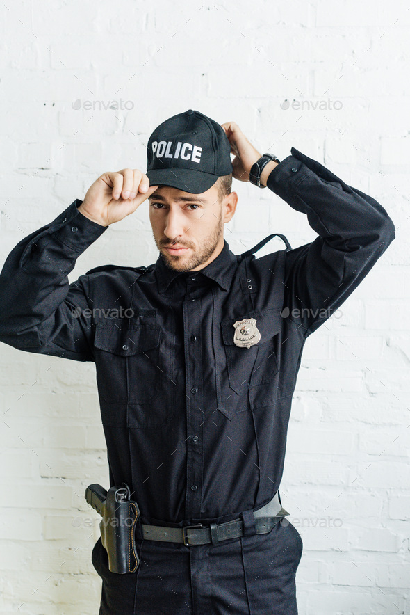 attractive young policeman putting on cap in front of white brick wall ...