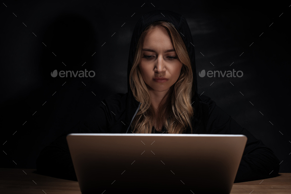 portrait of female hacker in black hoodie using laptop at wooden ...