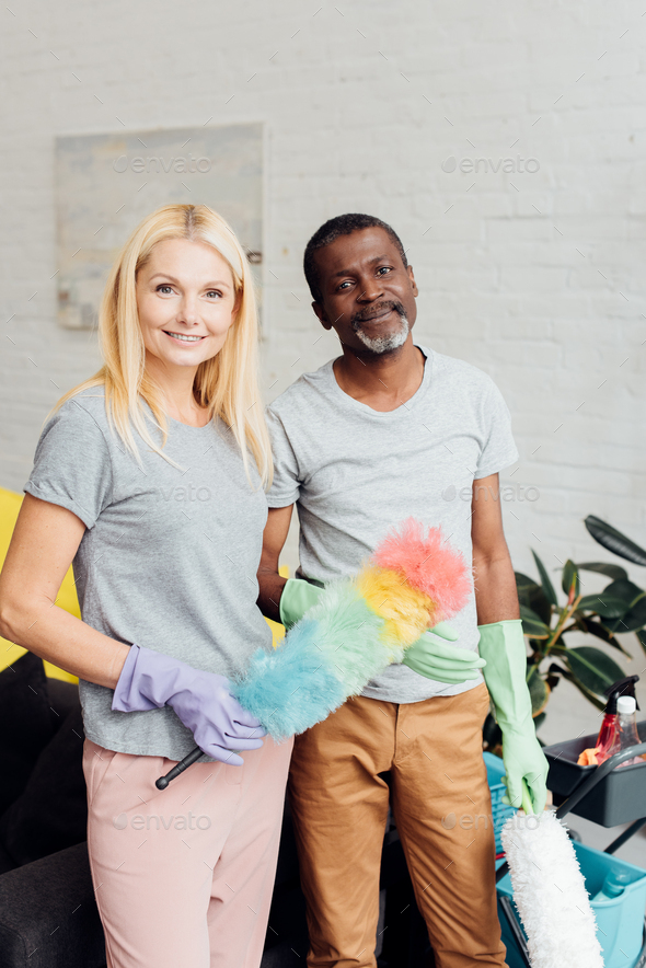 smiling blonde woman and african american man in rubber gloves holding dusters Stock Photo by ...