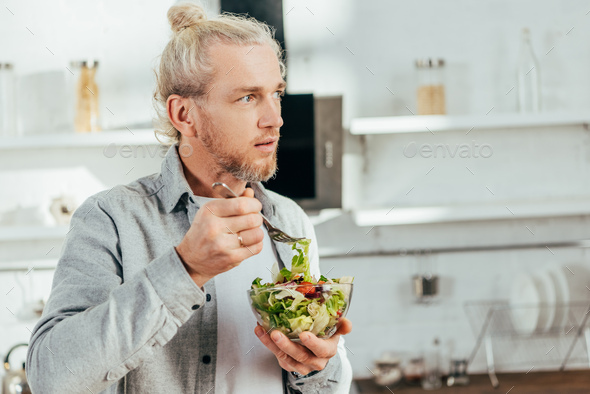 handsome man eating vegetable salad and looking away in kitchen Stock ...