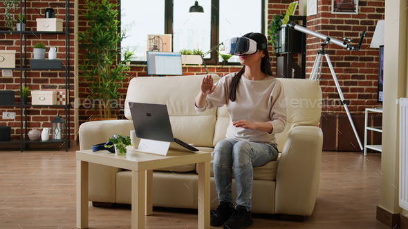 Playful woman with VR goggles playing on metaverse inside living room ...