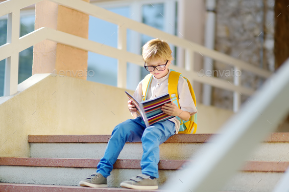 Smart little child sitting and reading on the stairs of school building ...