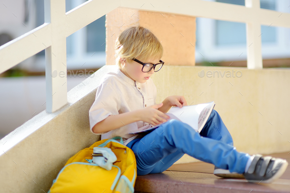 Smart little child sitting and reading on the stairs of school building ...