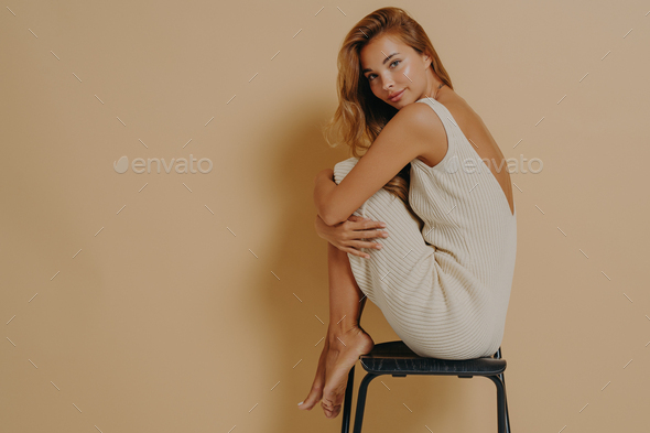 Beautiful young woman poses in profile on chair with legs curled up ...
