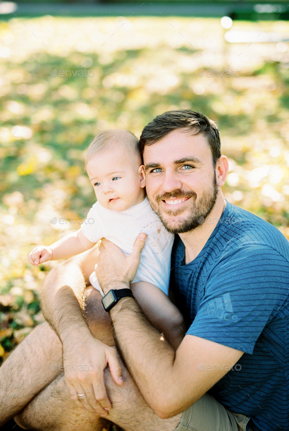 Smiling dad sitting with baby on lap in park Stock Photo by Nadtochii