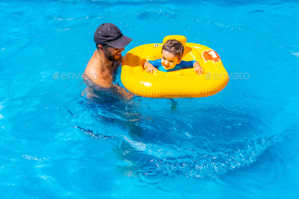 Smiling father playing with his son with a yellow float in the pool in ...
