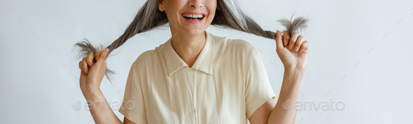 Happy middle aged Asian woman holds twisted hoary hair on light grey ...