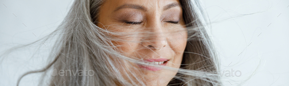 Happy mature Asian model with flying hoary hair stands on light grey ...