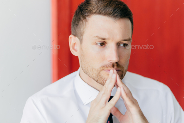 Thoughtful attractive male keeps hands together near mouth, has pensive ...