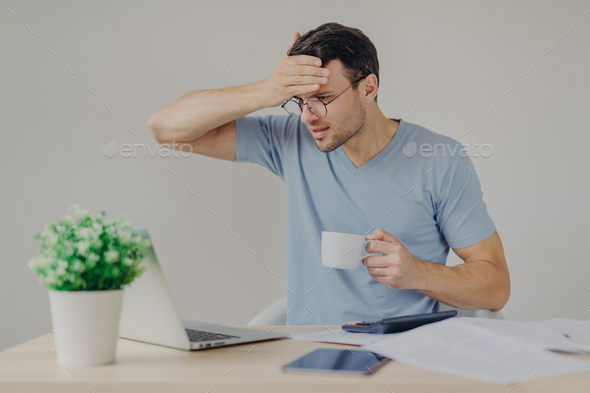 Troublesome brunet man in casual t shirt, has headache as calculates ...