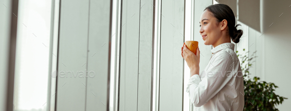 Smiling asian female worker drinking coffee standing near windows at ...