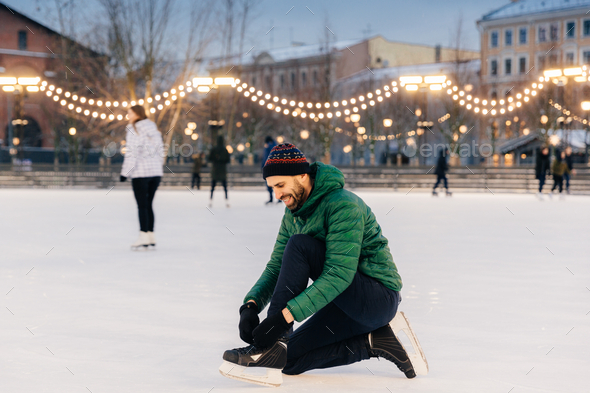 Portrait of cheerful male laces up skates as going to go skating on ice ...