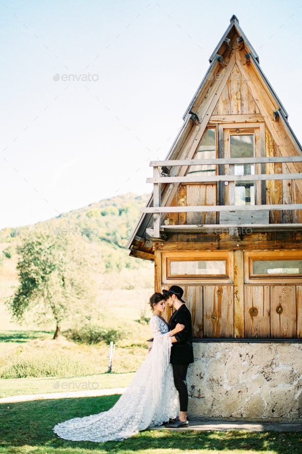Groom hugs bride in a white dress near the wall of a triangular house ...