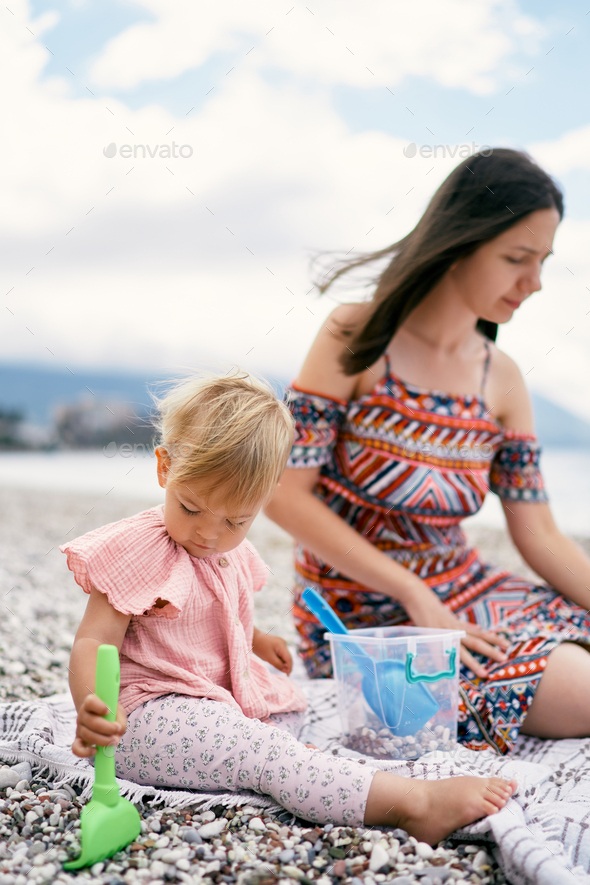 Little girl playing toy rake while sitting on a blanket on a pebble ...