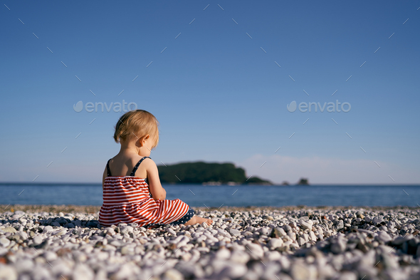 Little girl sits on a pebble beach with her head bowed. Back view Stock ...