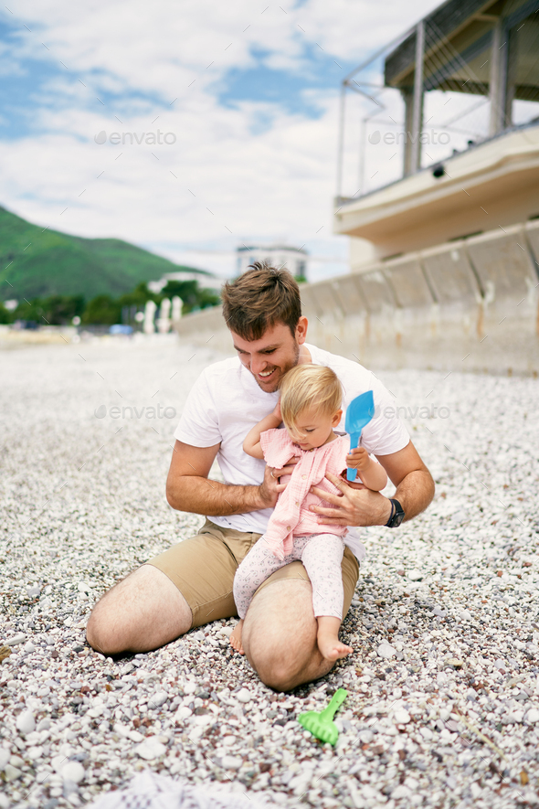 Dad holds little girl on his lap while sitting on pebble beach Stock ...