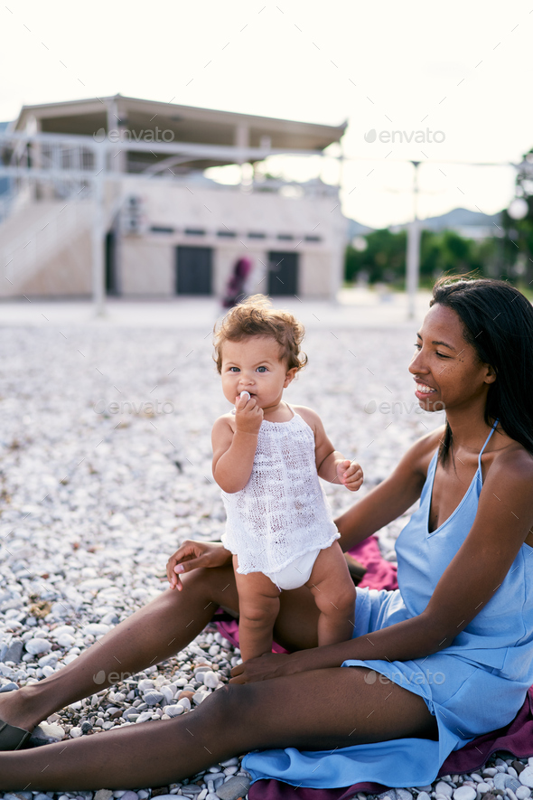 Little girl gnaws pebbles while standing next to sitting mother on the ...