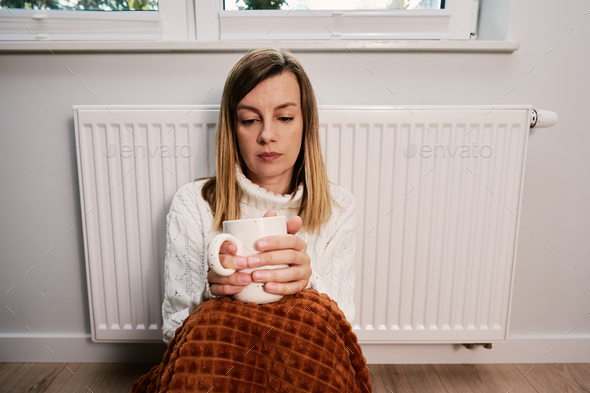 Worried woman sit near heating radiator under blanket with cup of tea ...