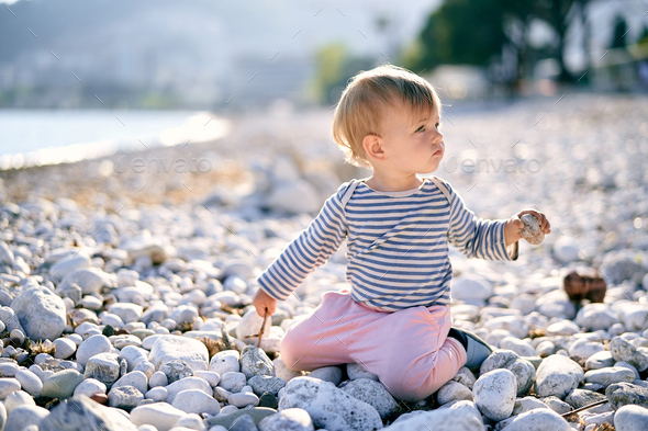 Kid sits on his knees with his head turned on a pebble beach and holds ...