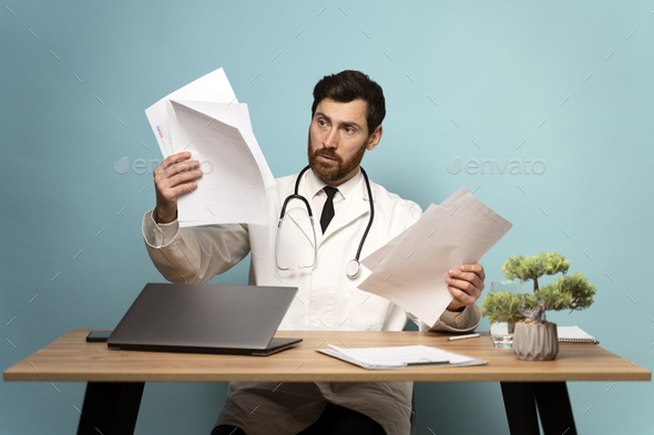 Serious doctor looking at his documents while sitting at his workplace ...