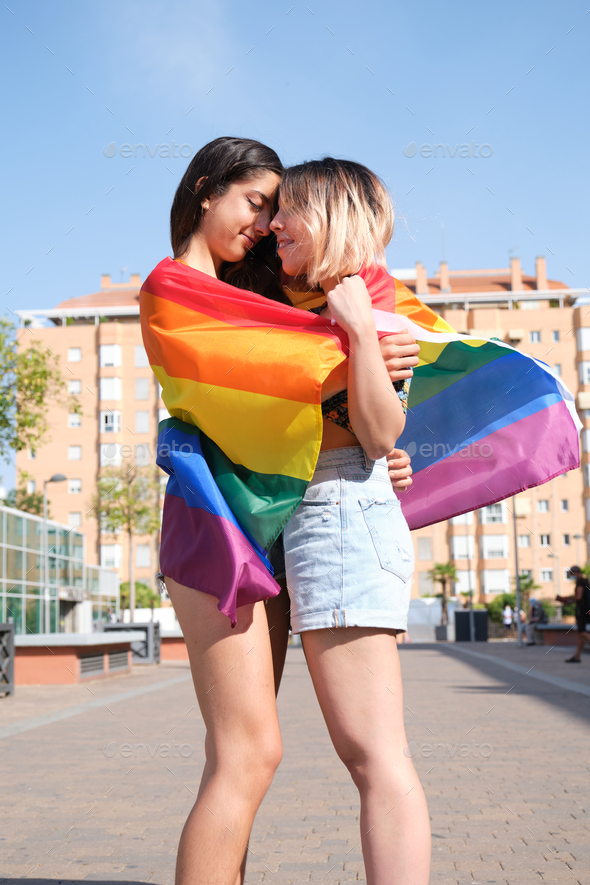 Young lesbian couple smiling and hugging wrapped in a rainbow flag ...