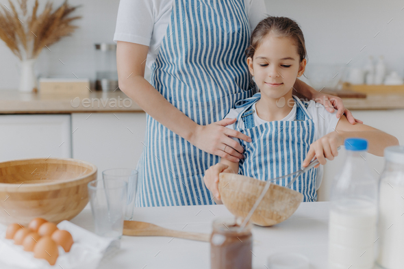 affectionate mother in apron embraces daughter who learns how to make ...