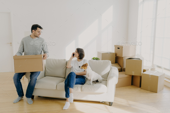 Photo of happy couple move in new home, pose on sofa with pet and boxes ...