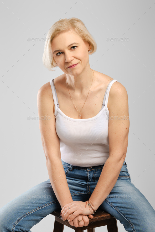 Barefoot senior woman in jeans and tank top sitting on chair in studio ...