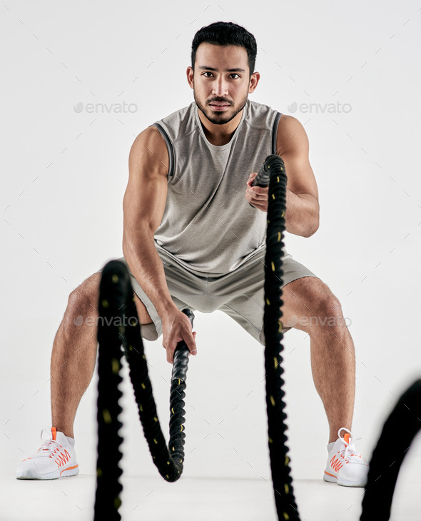 Studio portrait of a muscular young man exercising with battle ropes ...