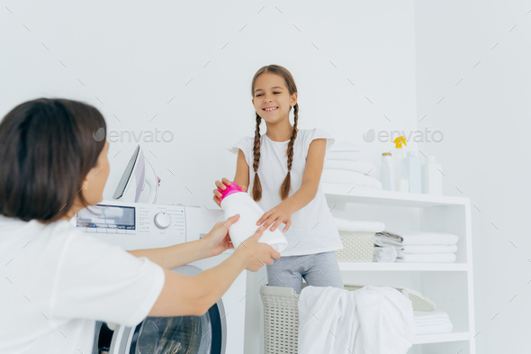 Cheerful girl gives washing detergent to mother, being in laundry room ...