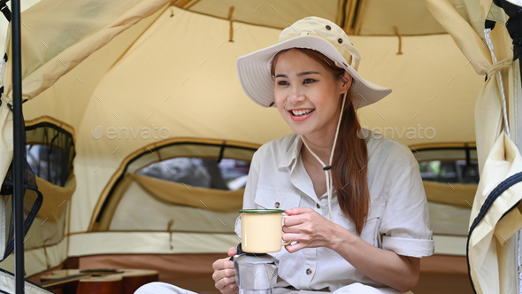 Pleasant asian woman sitting inside camp tent and drinking coffee ...