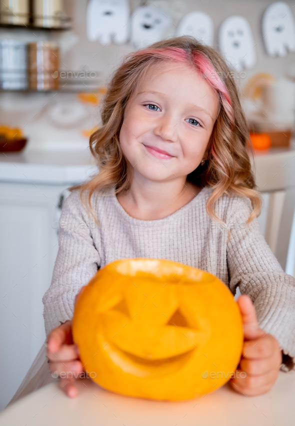 Cute little girl holding pumpkin with spooky face, sitting in kitchen ...