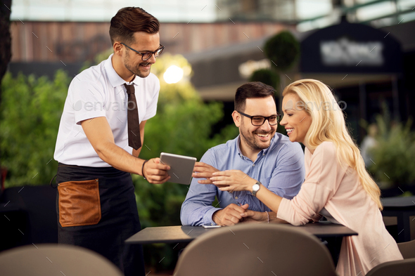 Happy waiter showing the menu on touchpad to his guests in a cafe ...
