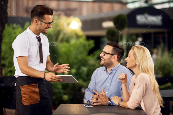 Happy waiter communicating with a couple while showing them menu on ...