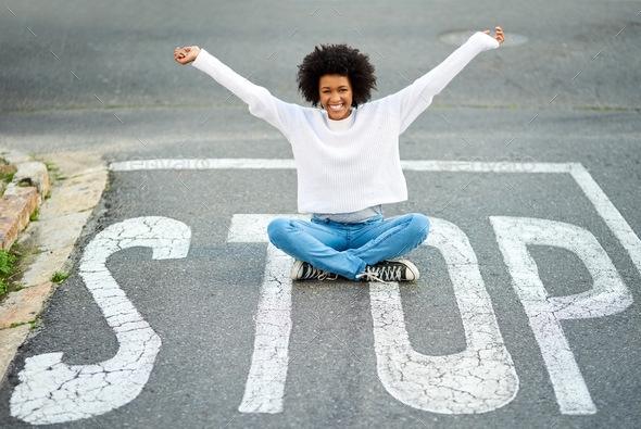 Shot of a happy young woman sitting on a stop sign at an intersection ...