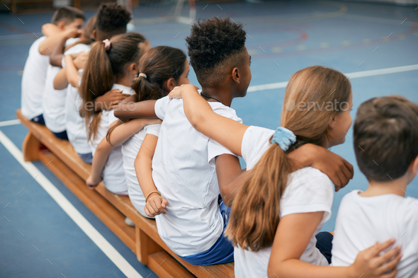 Rear view of large group of school children sitting embraced during PE ...