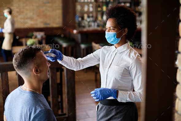 Black waitress measuring temperature of guest with infrared thermometer ...