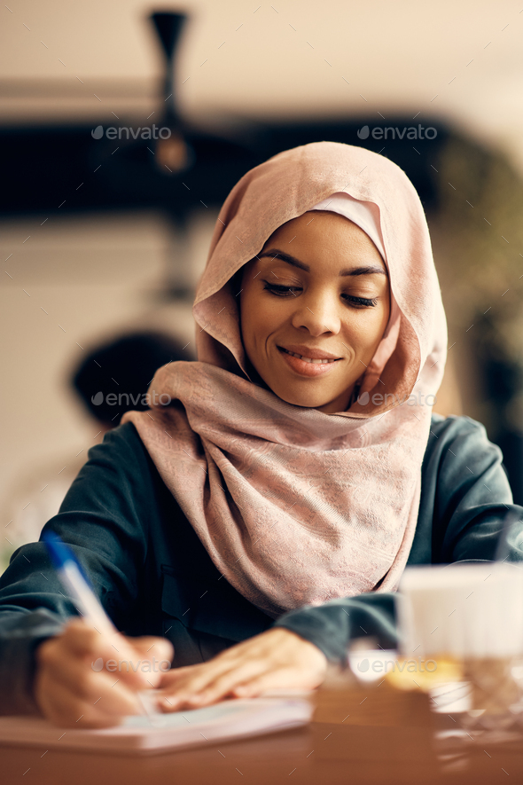 Smiling Muslim woman in hijab writing notes while studying in cafeteria ...