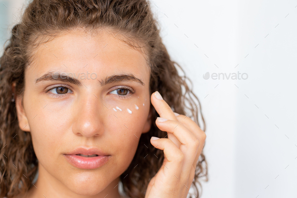 Beautiful young woman applying cosmetic cream treatment on her face isolated on white background ...