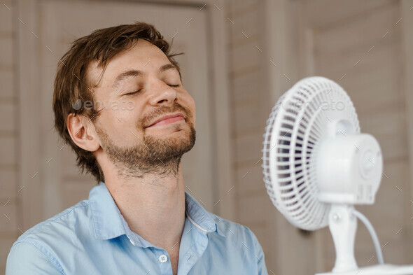 Sweaty man enjoying air flow from fan. Businessman refreshing in front ...