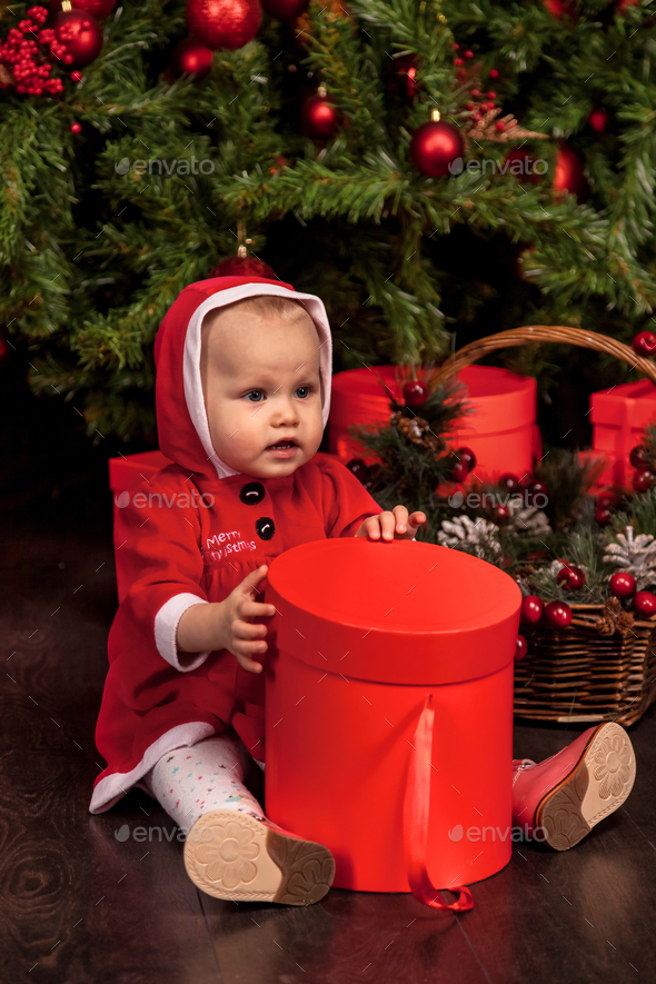 One-year-old girl disguised Santa Claus with gift sits at background of ...