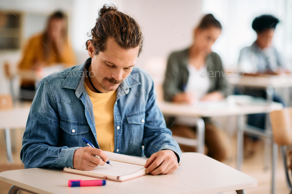 Male student writing notes while attending a lecture in the classroom ...