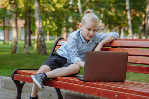 cute caucasian boy sitting on bench in park with laptop computer. Black ...